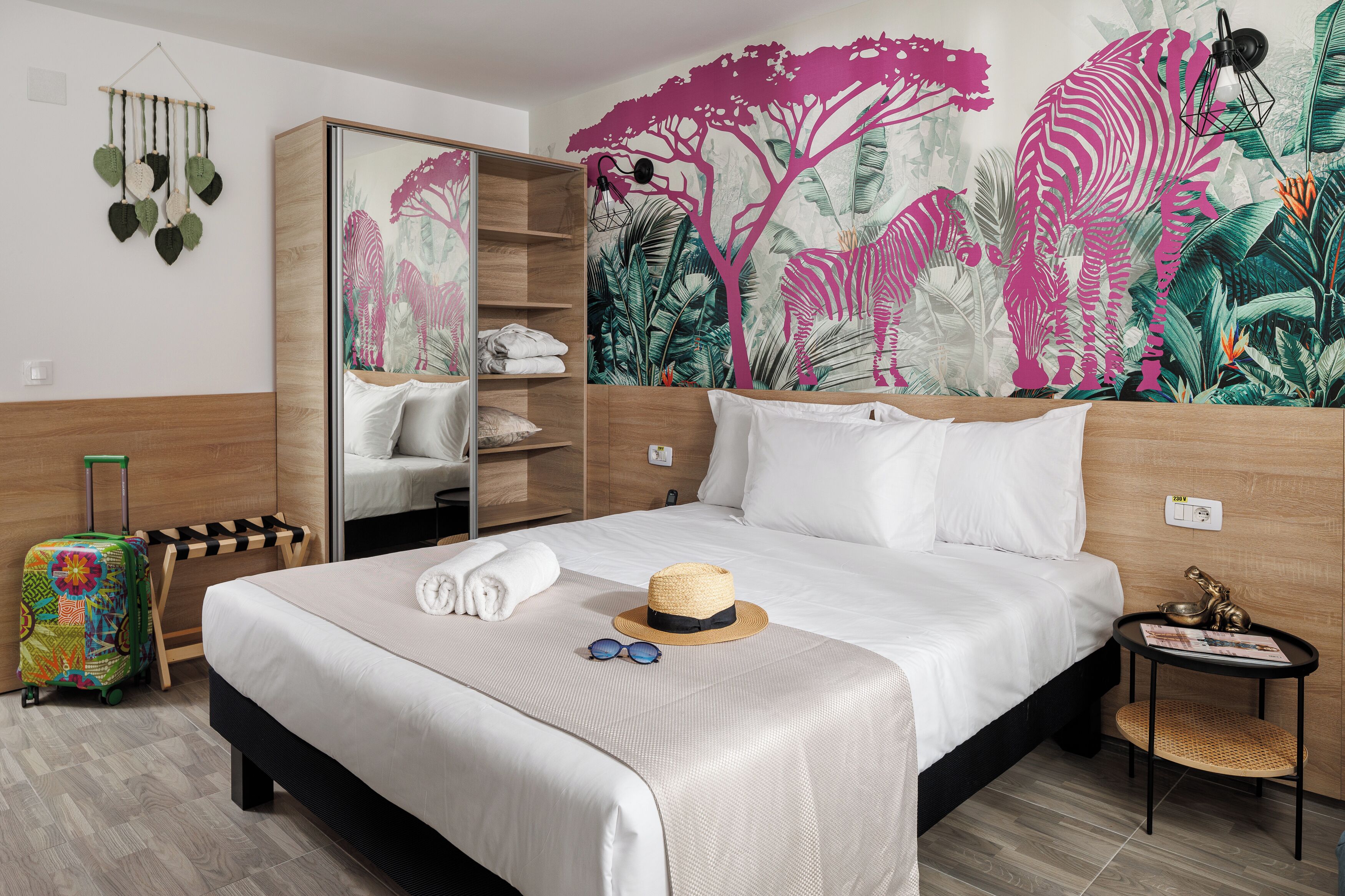 Cama bem arrumada com chapéu de palha e óculos de sol, uma mala e uma pequena mesa com abajur e objeto decorativo.