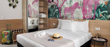 Cama bem arrumada com chapéu de palha e óculos de sol, uma mala e uma pequena mesa com abajur e objeto decorativo.