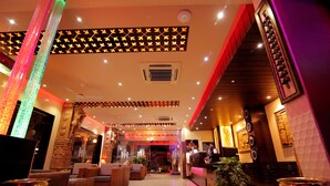 Um saguão de hotel com balcão de recepção, área de estar e elementos decorativos, como uma planta grande e luminárias fixadas na parede.