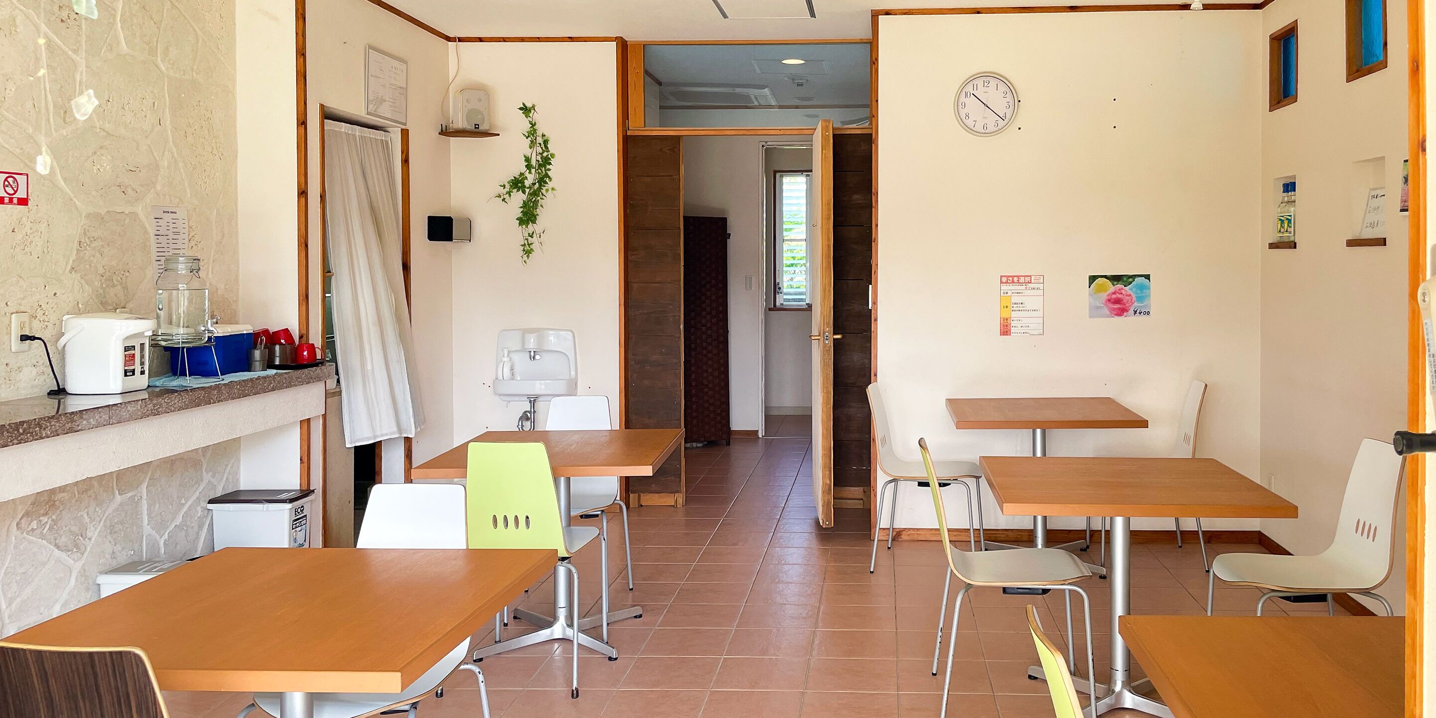 Quarto de hotel com cozinha compacta, área de refeições com mesas e cadeiras, relógio na parede e planta na parede.