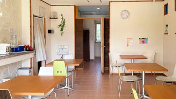 Quarto de hotel com cozinha compacta, área de refeições com mesas e cadeiras, relógio na parede e planta na parede.