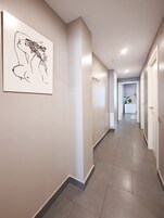 Um corredor moderno com design minimalista, apresentando uma grande obra de arte abstrata na parede esquerda, piso limpo e uma porta que dá para um cômodo com uma planta.
