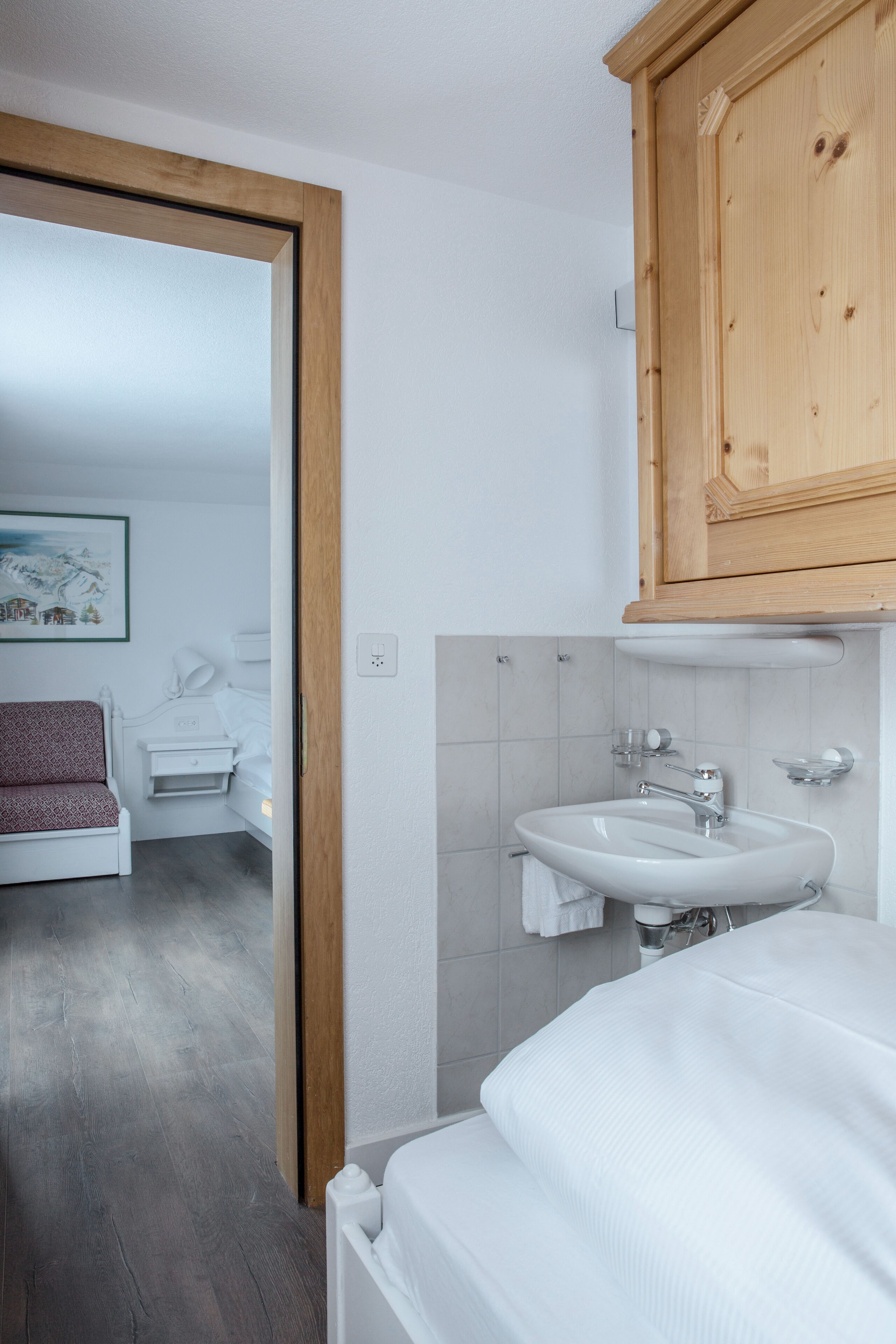 Casa de banho moderna com lavatório branco, armário de madeira e cama com roupa de cama branca.