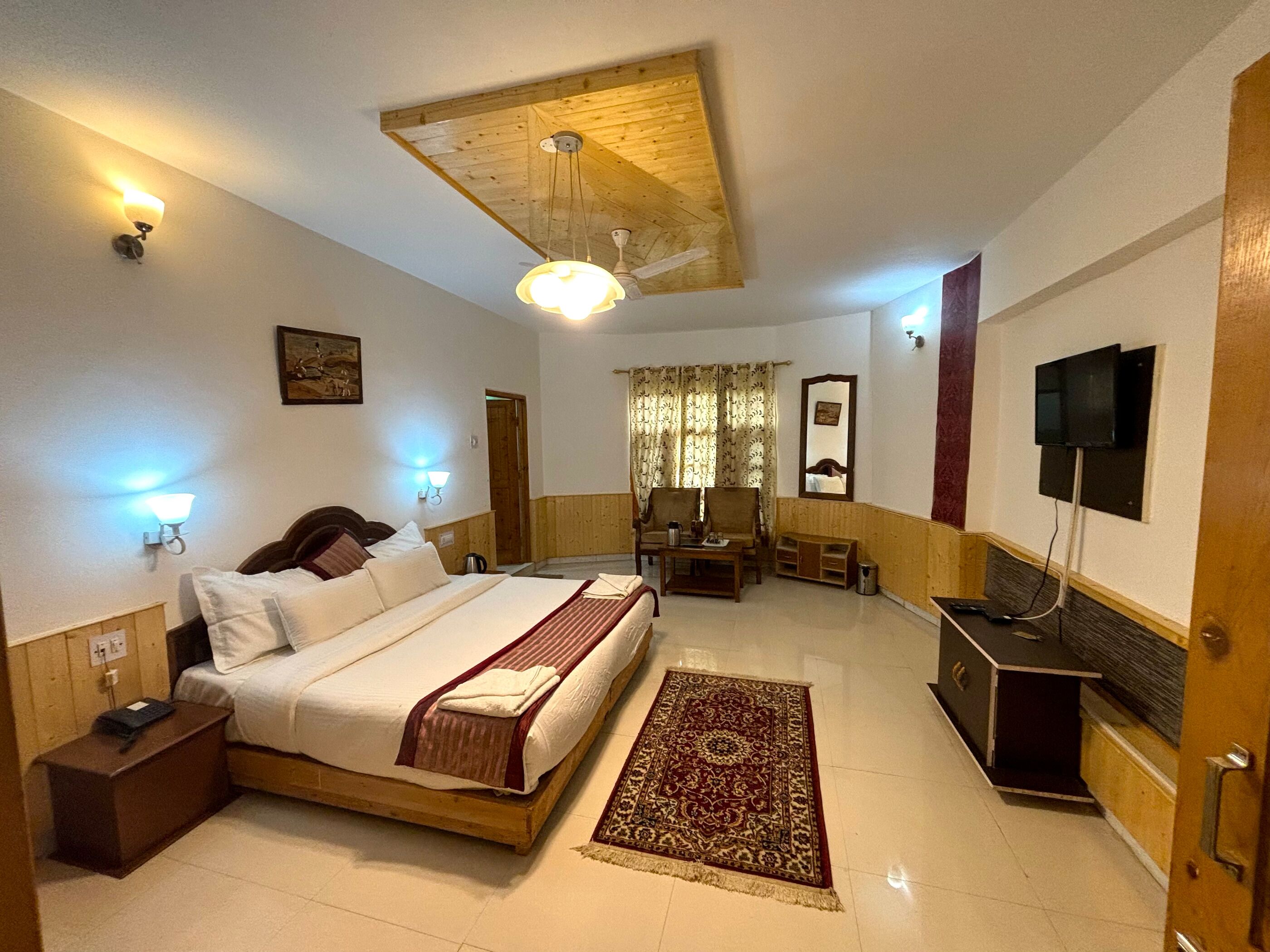 Quarto de hotel com teto de madeira, cama com lençóis brancos, área de estar com poltrona e mesa, televisão na parede e um espelho grande.