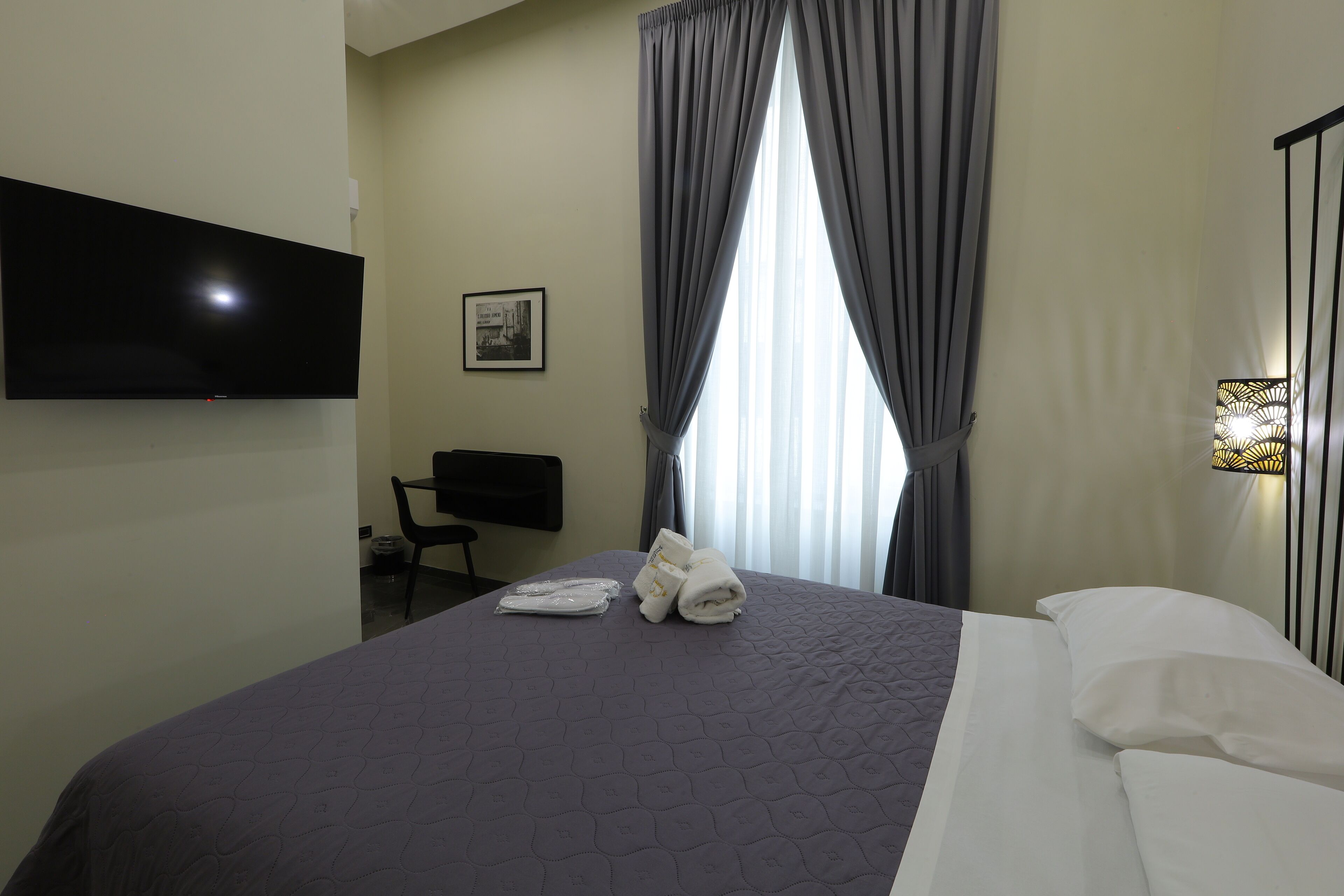 Quarto de hotel com uma cama grande, televisão de tela plana na parede, escrivaninha com cadeira e janela com cortinas.