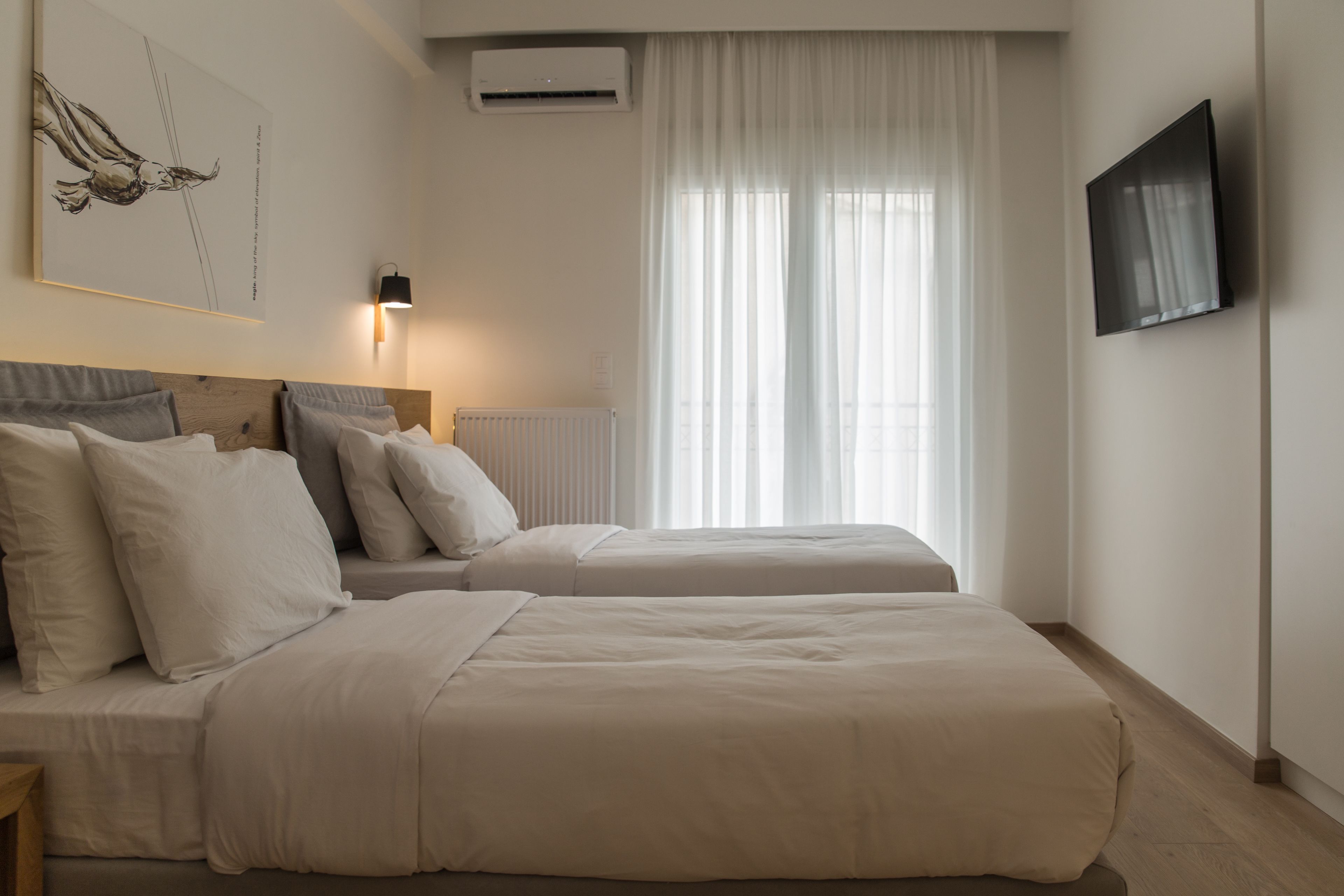 Quarto de hotel com duas camas, televisão presa à parede, radiador e uma janela grande com cortinas leves.