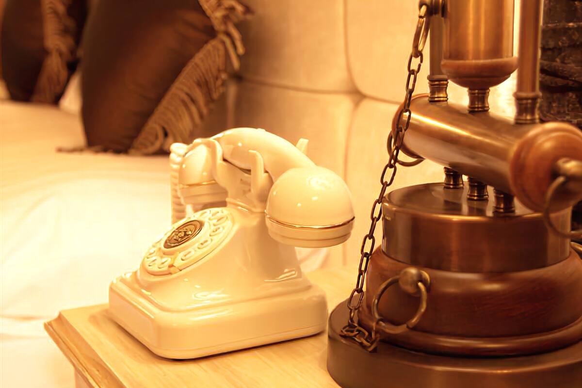 Um telefone rotativo antigo e uma cafeteira de latão sobre uma mesa de cabeceira de madeira em quarto de hotel.
