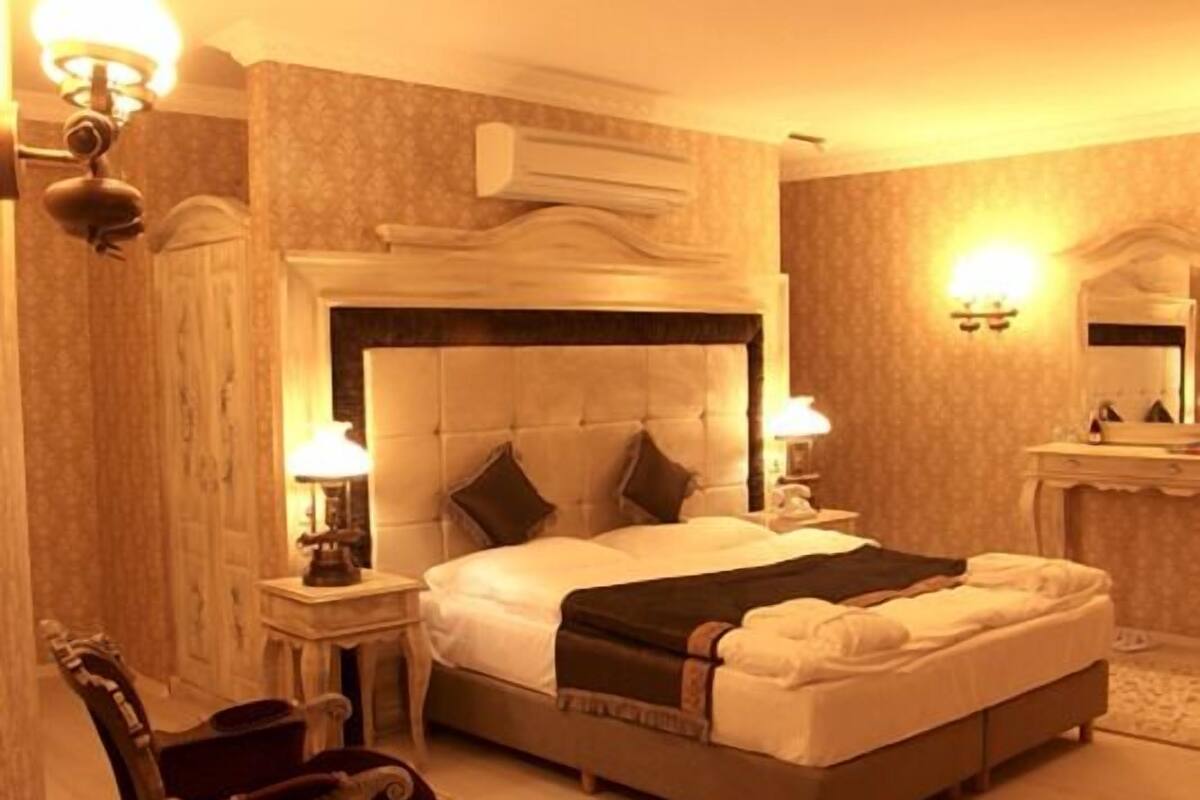 Quarto de hotel com uma cama grande, duas mesas de cabeceira com luminárias, uma cadeira, cômoda e espelho.