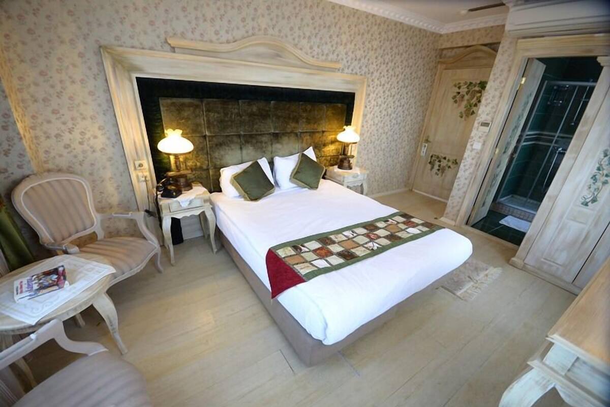 Quarto de hotel com uma cama grande, duas luminárias laterais, uma cadeira, uma mesinha de centro com uma revista e um banheiro visível através de uma porta de vidro.