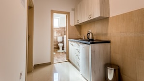 Uma cozinha compacta com forno embutido, pia e lixeira. Um armário de madeira fica acima do balcão, e uma porta dá acesso a um banheiro com vaso sanitário e espelho.