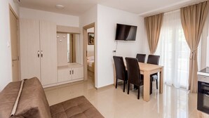 Sala de estar moderna com sofá, mesa de jantar com cadeiras, televisão na parede e área de cozinha com fogão.