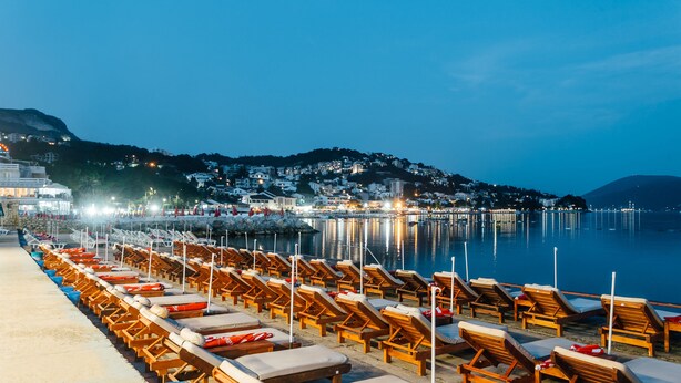 Um resort à beira-mar com fileiras de espreguiçadeiras de madeira voltadas para uma marina calma, um cenário montanhoso e edifícios com luzes refletindo na água.