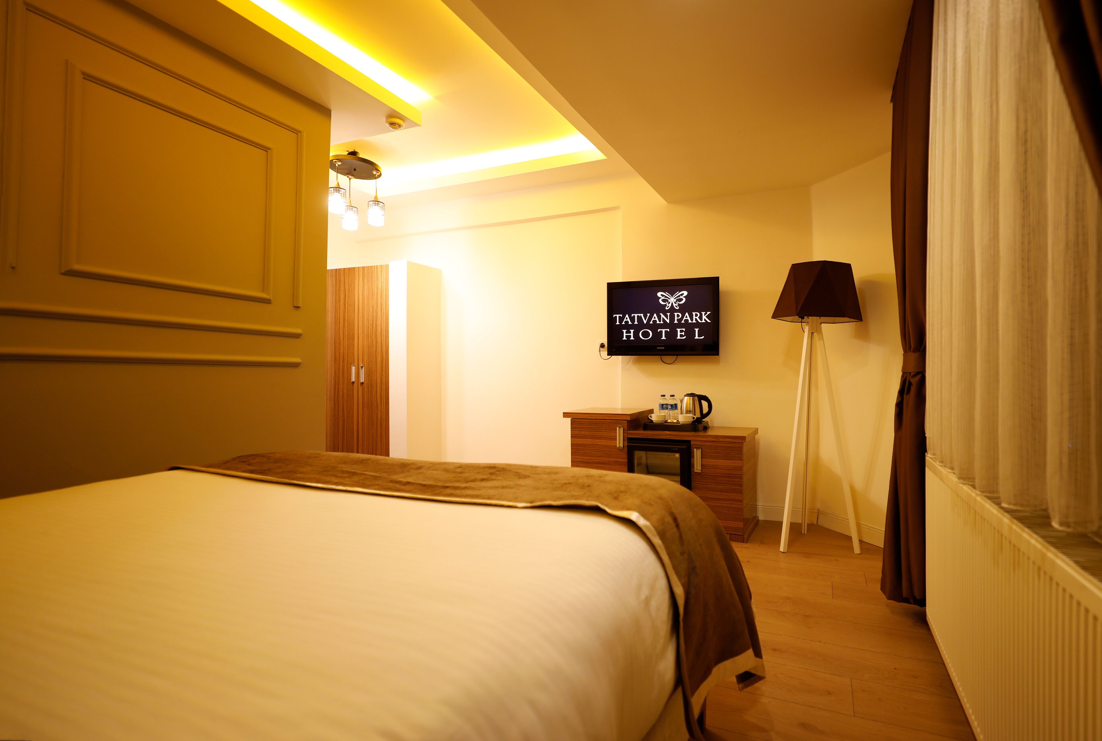 Quarto de hotel com cama, televisão exibindo o nome do hotel, escrivaninha com abajur e janela com cortinas.
