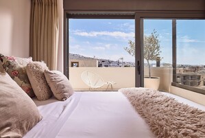Quarto com cama grande, tapete macio, uma cadeira e vista da cidade através de uma janela ampla.
