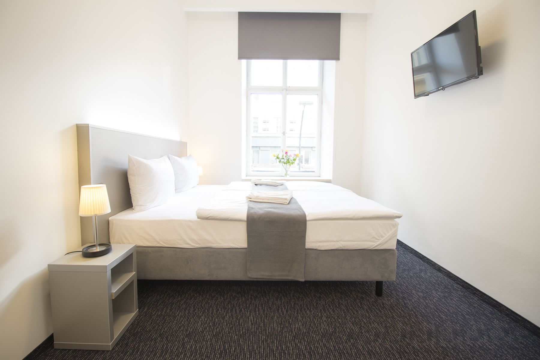 Quarto de hotel com cama, mesa de cabeceira com abajur, TV na parede e janela com vista para edifícios.