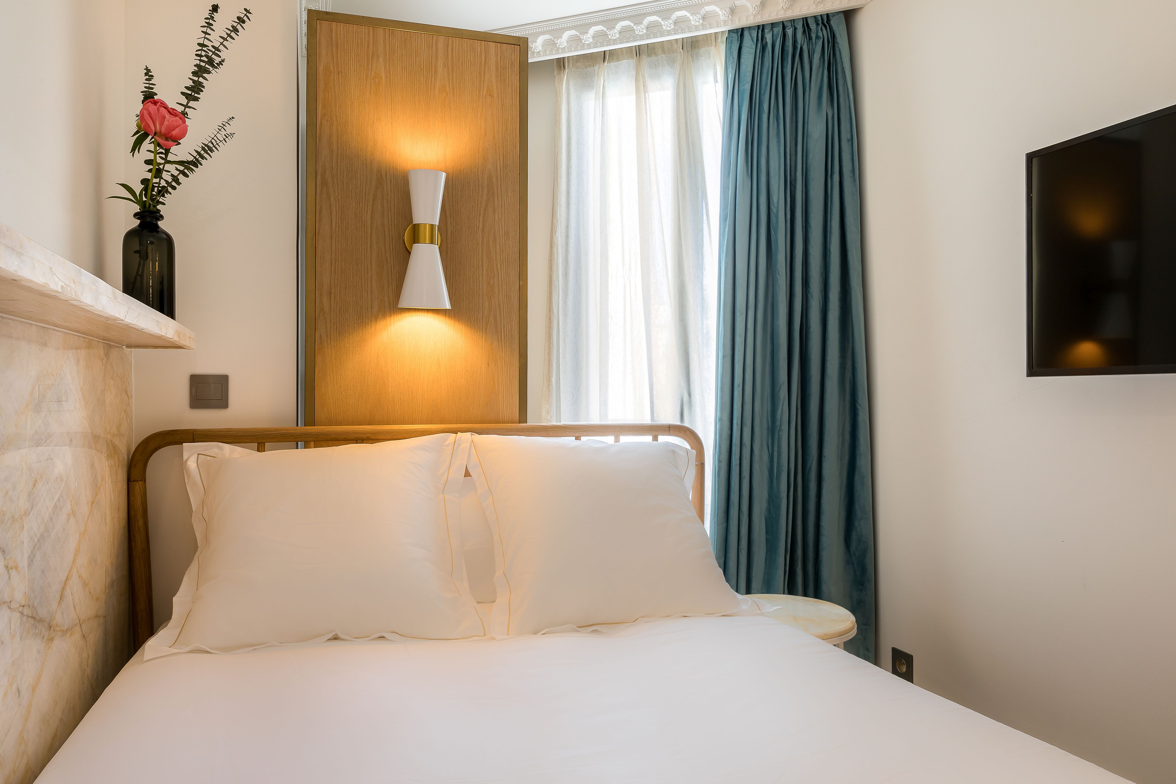 Quarto de hotel com cama, um vaso com flor, luminária na parede, janela com cortinas e televisão na parede.