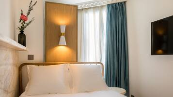 Quarto de hotel com cama, um vaso com flor, luminária na parede, janela com cortinas e televisão na parede.