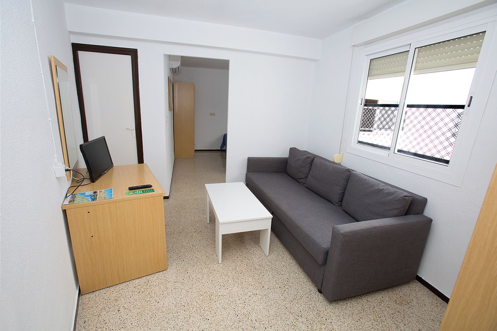 Quarto pequeno e pouco mobiliado, com uma escrivaninha de madeira, uma mesa branca e um sofá cinzento. Há uma janela com persianas, uma porta que dá para outro espaço e um espelho na parede.