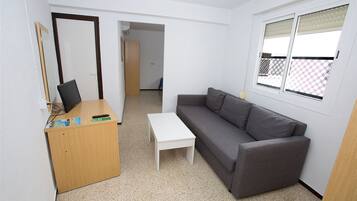 Quarto pequeno e pouco mobiliado, com uma escrivaninha de madeira, uma mesa branca e um sofá cinzento. Há uma janela com persianas, uma porta que dá para outro espaço e um espelho na parede.