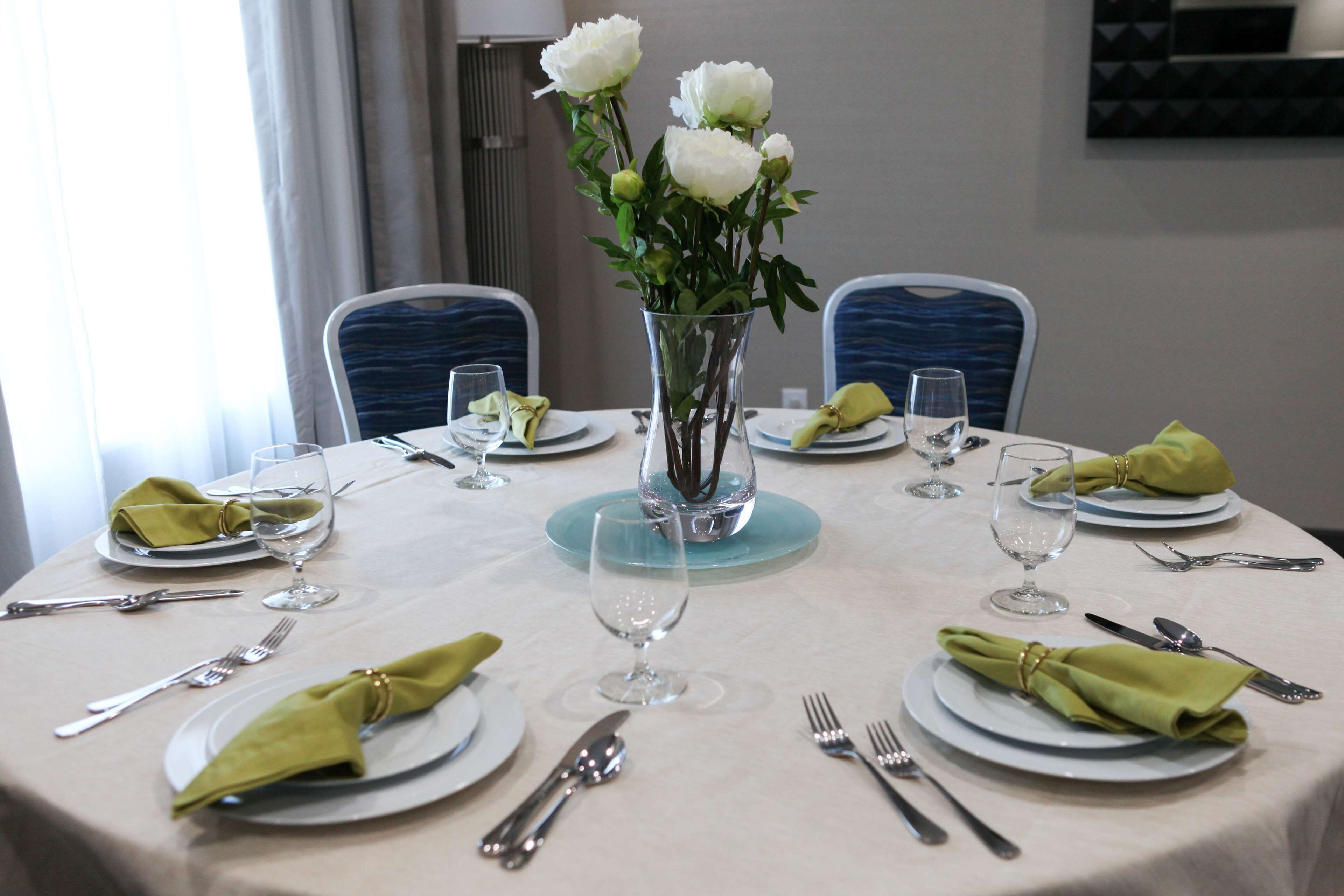 Mesa de jantar posta para quatro com pratos brancos, guardanapos verdes e um vaso transparente com flores brancas.