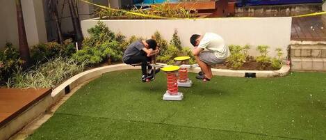 Duas pessoas jogando um jogo em grama sintética, com piscina e edifícios ao fundo.