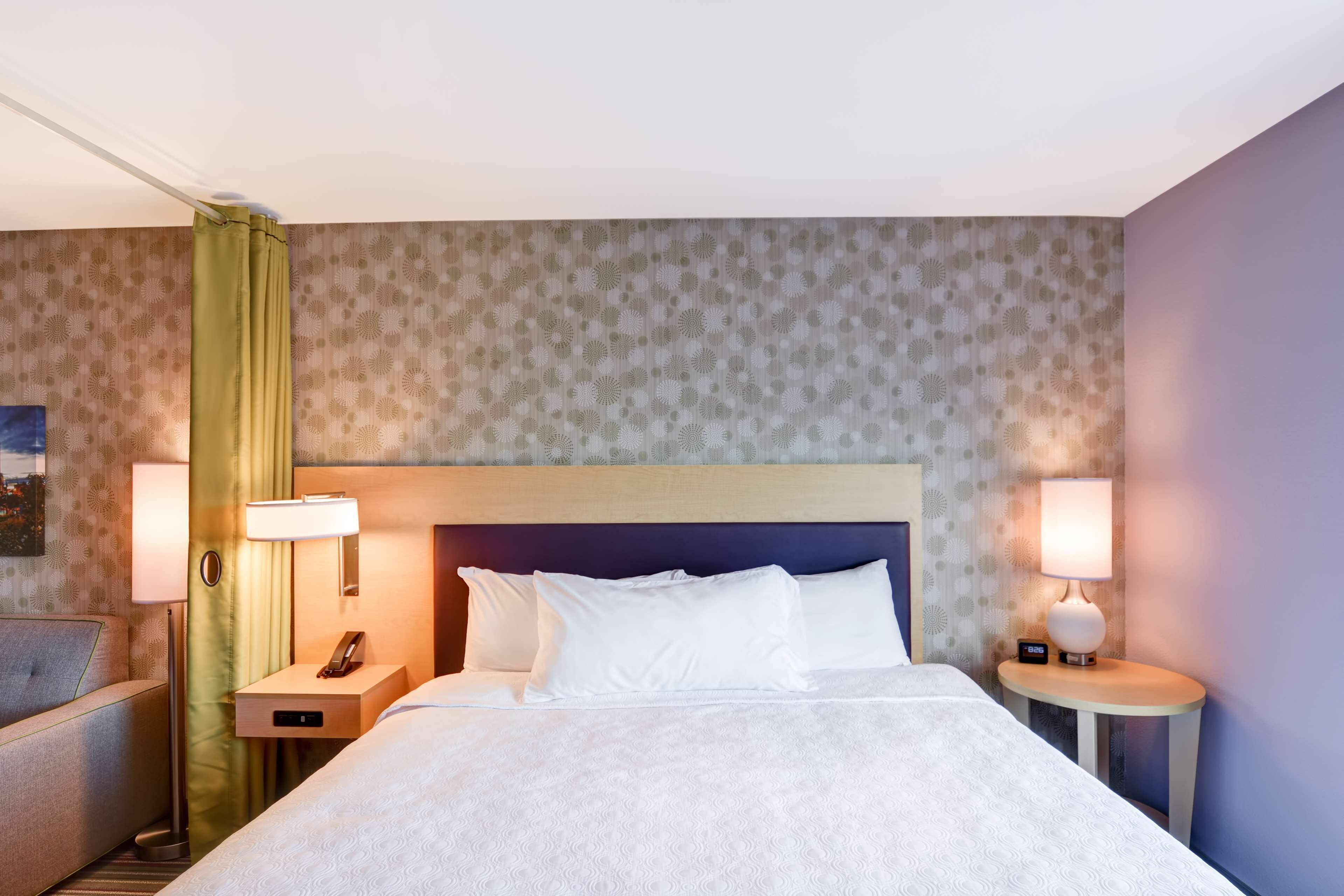 Quarto de hotel com cama bem arrumada, duas mesas de cabeceira com luminárias, uma cadeira e papel de parede estampado.