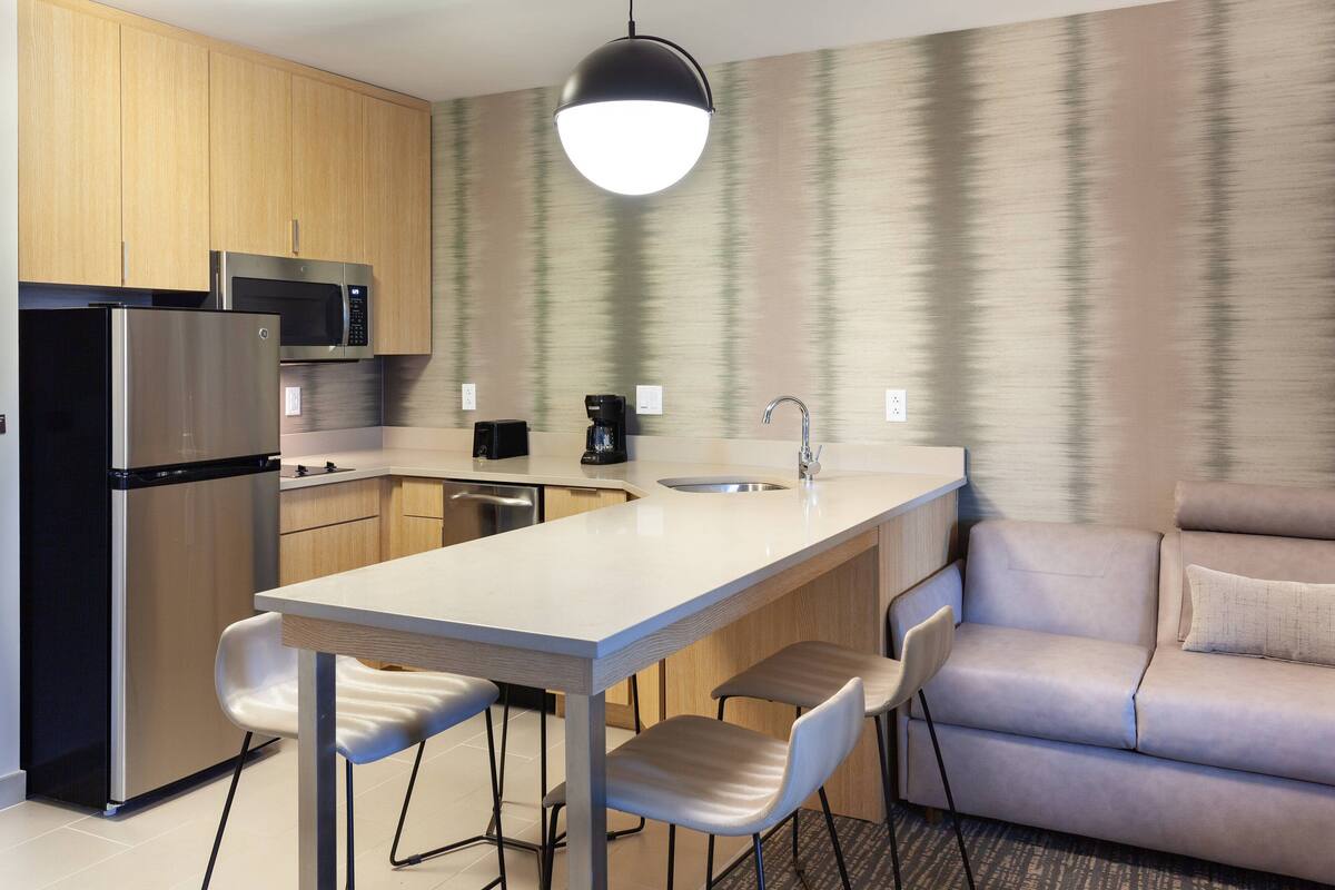 Uma cozinha compacta com geladeira, micro-ondas e cafeteira, uma mesa de jantar branca com cadeiras, e um sofá na área de estar adjacente.