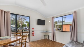 Um quarto com uma mesa de jantar de madeira, cadeiras, um sofá, uma televisão fixada na parede e janelas amplas com cortinas.