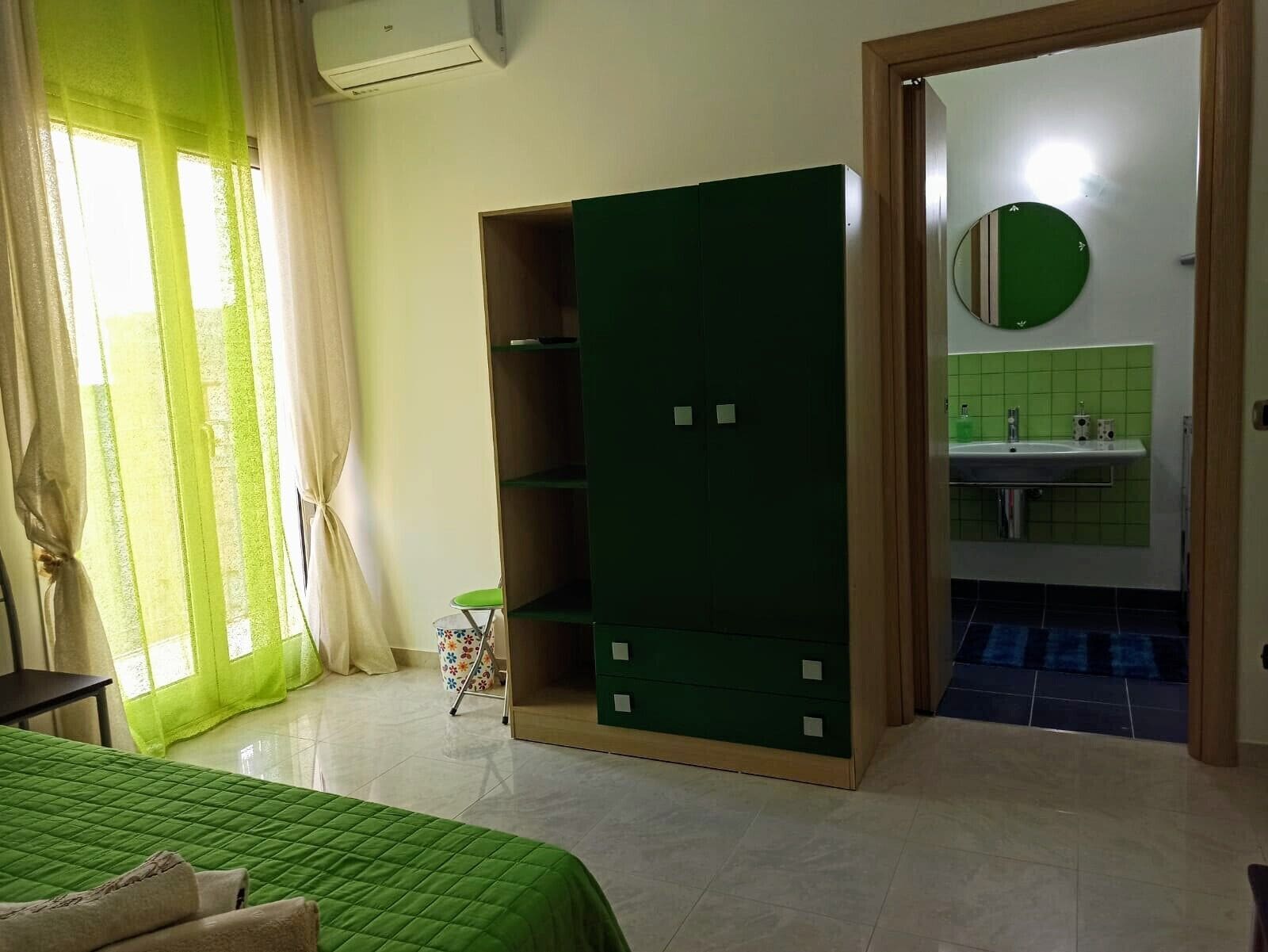 Quarto com cama, guarda-roupa, um banquinho pequeno e banheiro com pia e espelho.