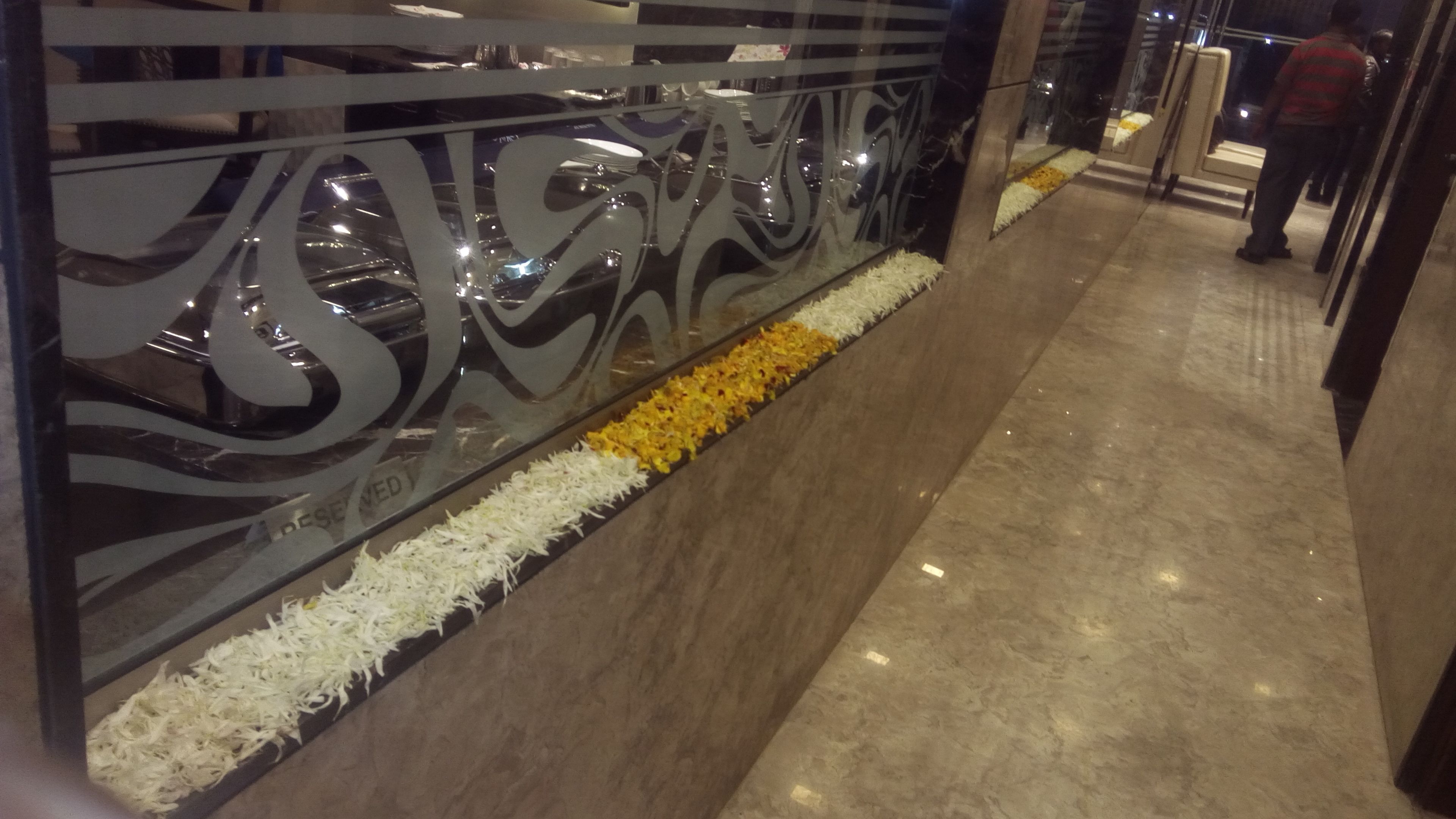 Uma área de recepção com design moderno, apresentando um painel de vidro com padrões decorativos, piso de mármore e um caminho delimitado por flores brancas e amarelas.