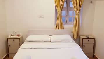 Cama bem arrumada com lençóis brancos, janela com cortinas amarelas, ventilador de teto e mesa de cabeceira pequena com abajur.