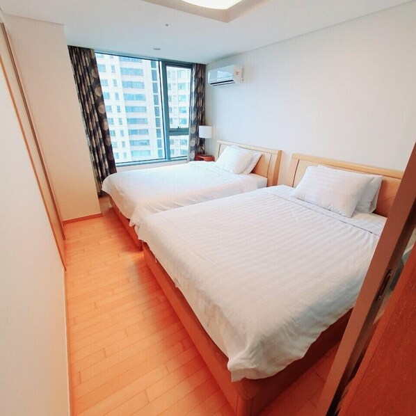 Quarto de hotel com duas camas, janela com cortinas, ar condicionado e piso de madeira.