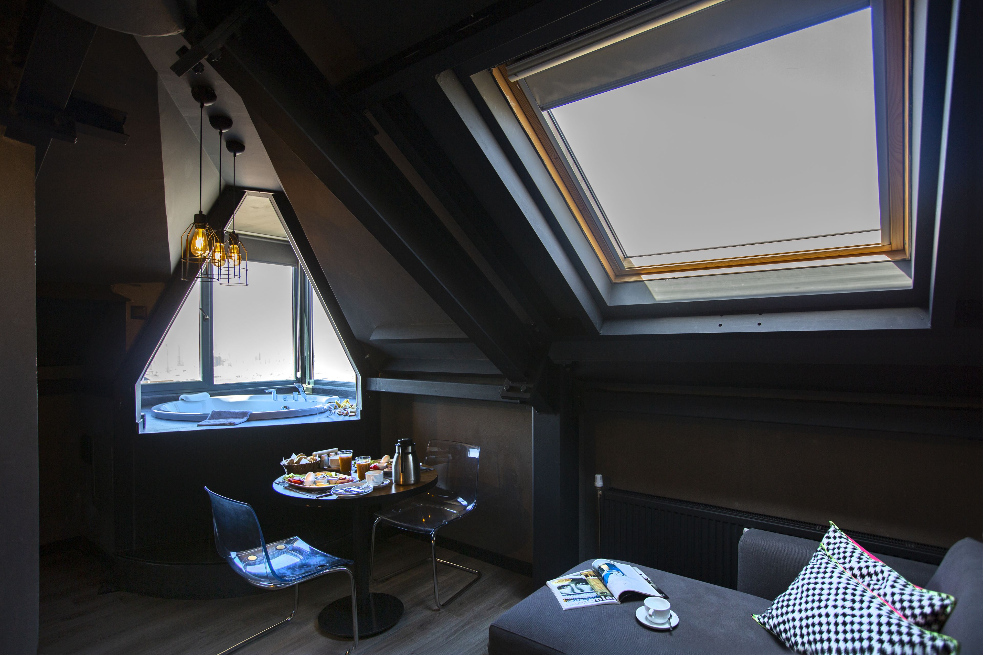 Um quarto moderno no estilo loft, com teto inclinado, claraboia, área de jantar com mesa e cadeiras, e um espaço de estar aconchegante com sofá e almofadas.