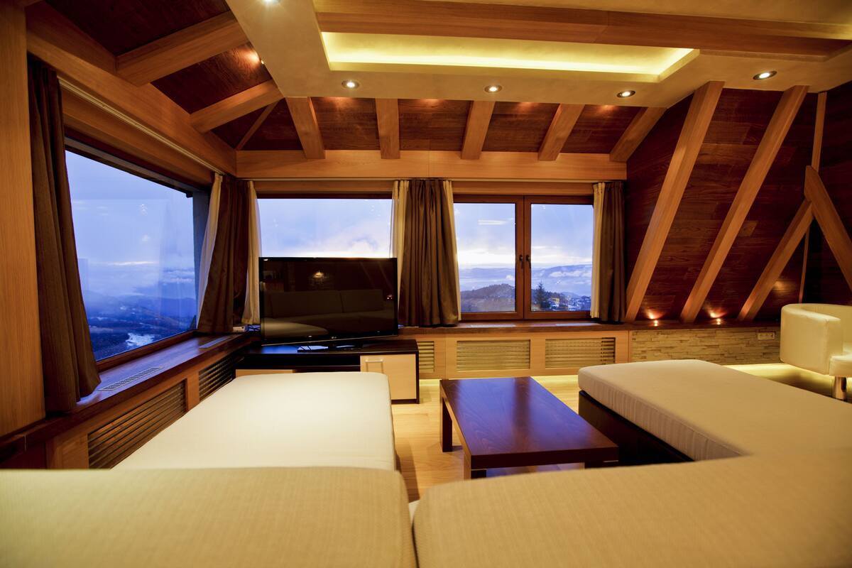 Quarto de hotel moderno com televisão grande, vigas de madeira, sofá, cama e vista para as montanhas.