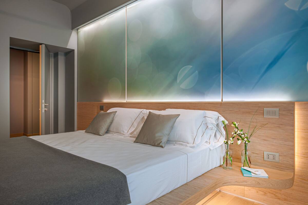 Quarto de hotel moderno com uma cama bem arrumada, uma mesa de cabeceira com um livro e um vaso de flores, e uma grande janela com vista para a cidade.