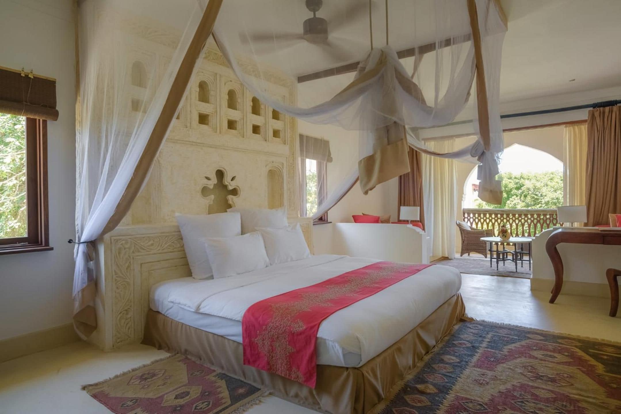 Quarto com cama grande, dossel, varanda com mesa e cadeiras, e vista para o verde lá fora.