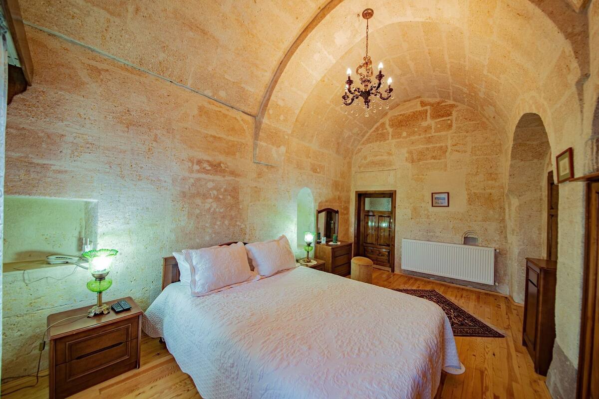 Quarto com uma cama grande, mesas de cabeceira de madeira, um candelabro e paredes de pedra.