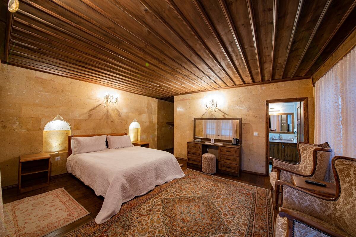 Quarto com teto de madeira, cama com colcha branca, mesa de cabeceira com abajur, cômoda, espelho e tapete estampado.