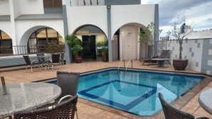 Uma piscina com espreguiçadeiras e mesas, rodeada por plantas em vasos e um edifício com janelas em arco.
