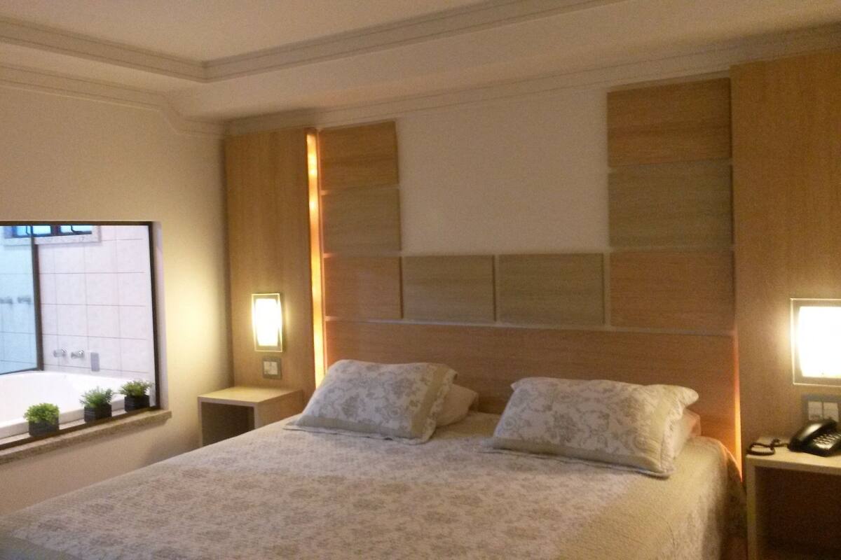 Quarto de hotel com uma cama grande, duas mesas de cabeceira, uma janela com vista e um candeeiro fixado na parede.