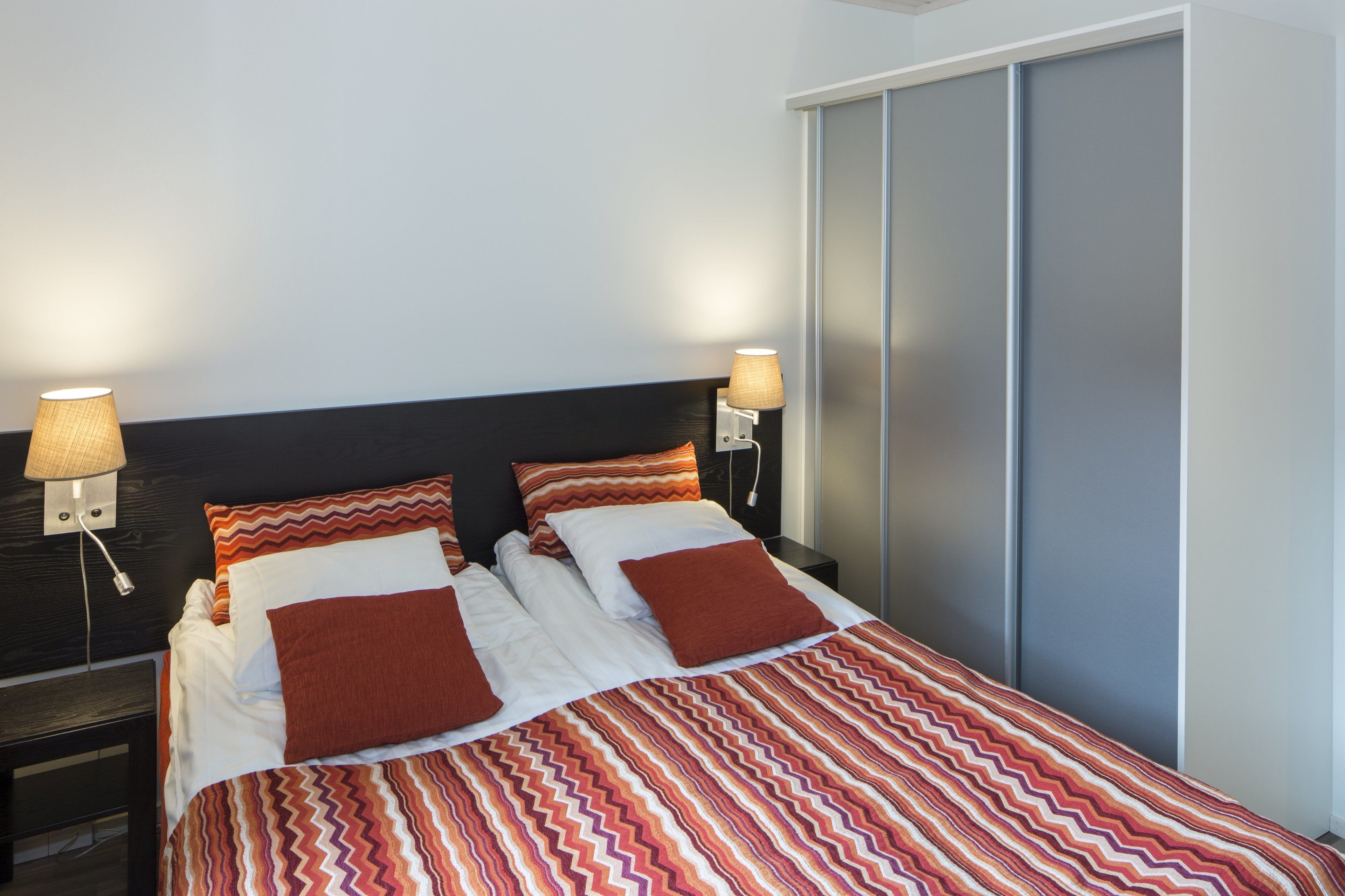 Cama bem arrumada com roupa de cama estampada, duas luminárias de cabeceira e uma porta de guarda-roupa deslizante.