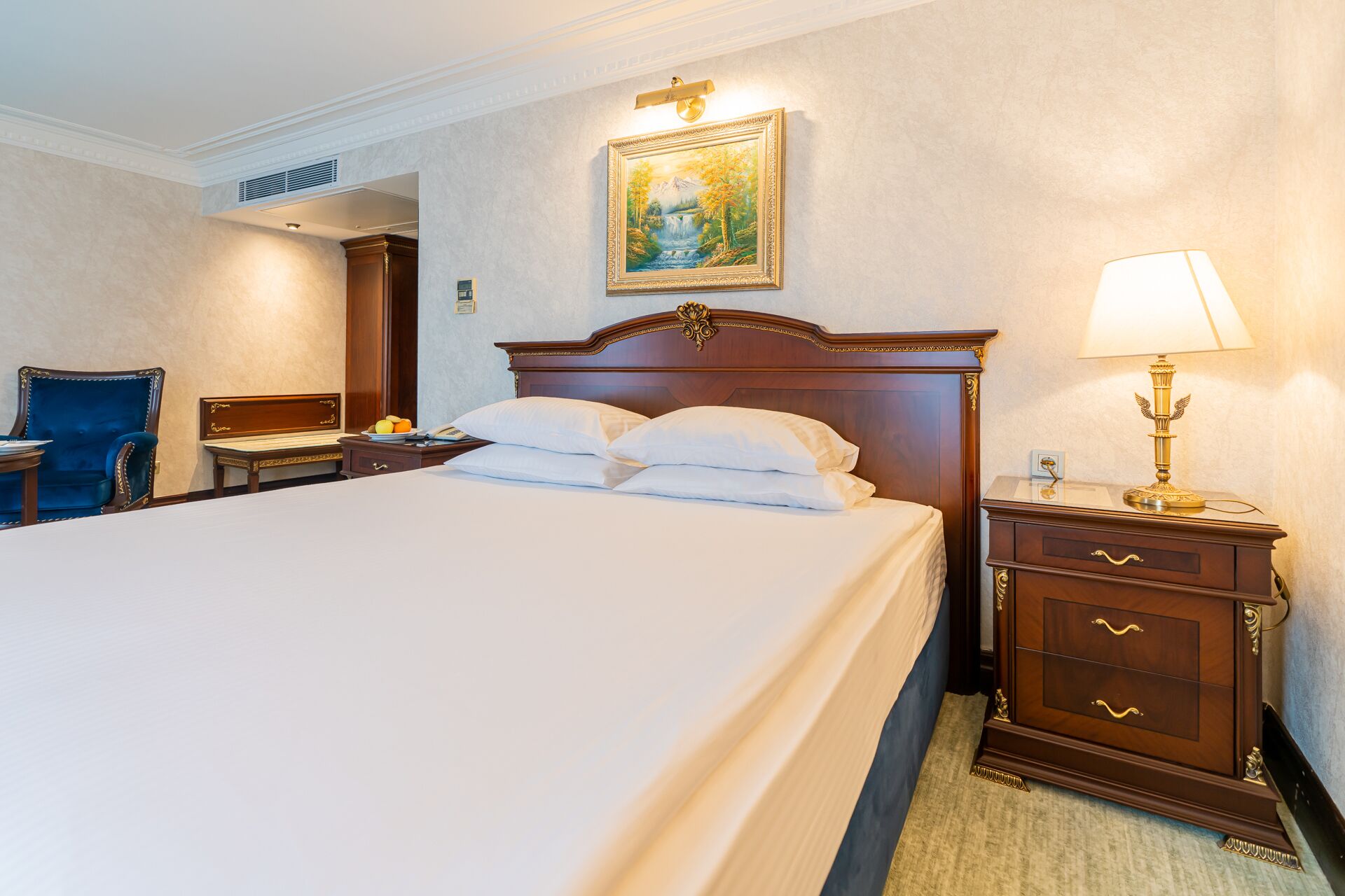 Quarto de hotel com uma cama grande, uma mesa de cabeceira com abajur, uma cadeira e um quadro na parede.