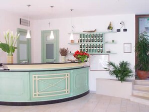 Uma área de recepção com balcão curvo, iluminação pendente, porta verde e prateleira com plantas na parede.