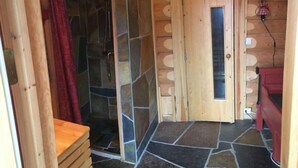 Interior de uma cabana de madeira com soalho de tijolos, área de duche com porta de vidro e um banco vermelho.
