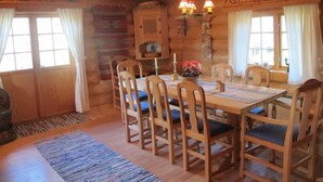 Interior de uma cabana de madeira com uma mesa de jantar rodeada por cadeiras, um lustre e um tapete.
