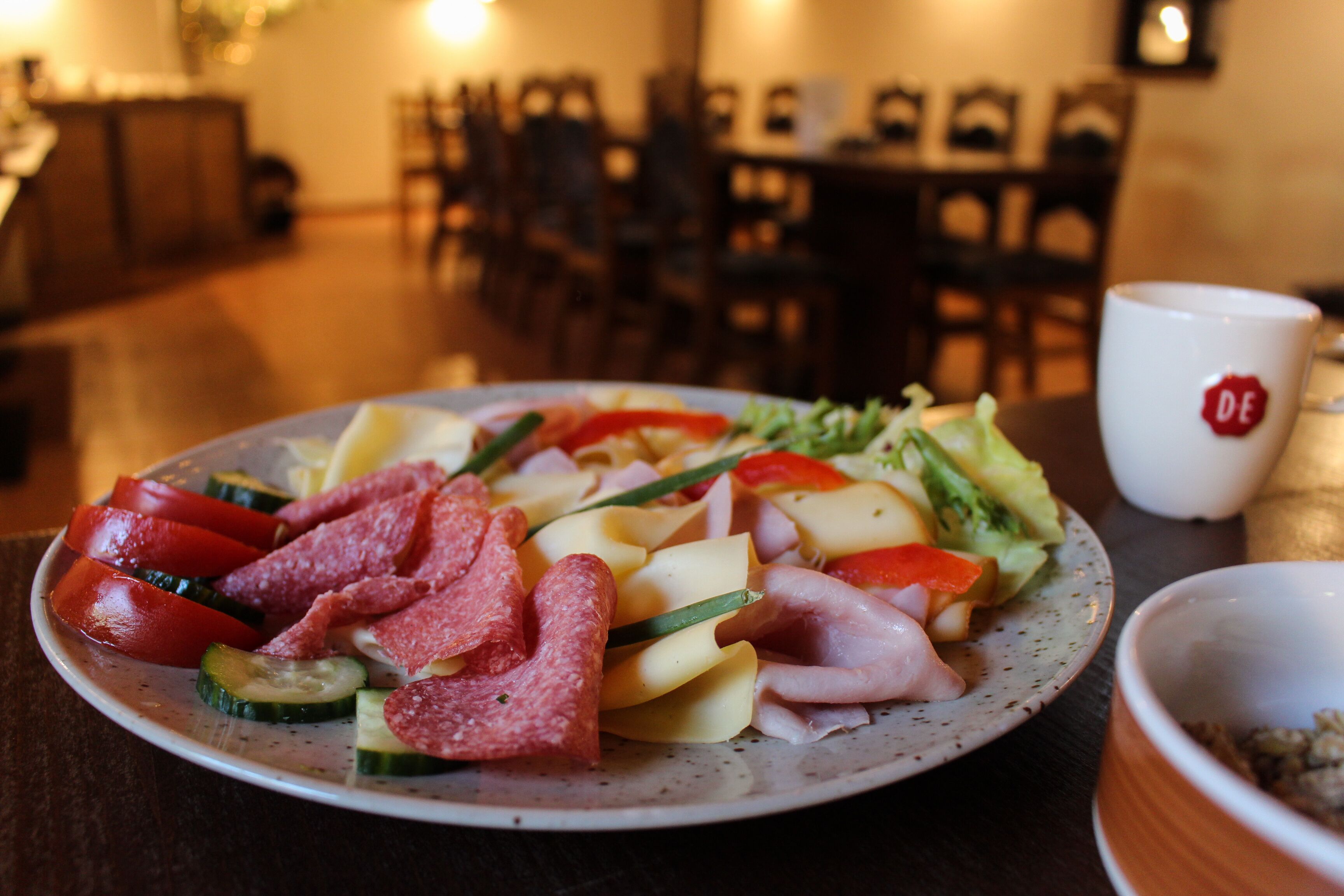 Um prato com legumes e carnes fatiadas sobre uma mesa em uma área de refeições, com cadeiras e outra mesa ao fundo.