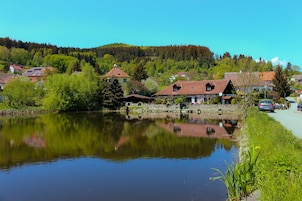 Uma aldeia tranquila à beira do lago, com casas tradicionais, um lago calmo refletindo a paisagem, e um caminho ao longo da margem.