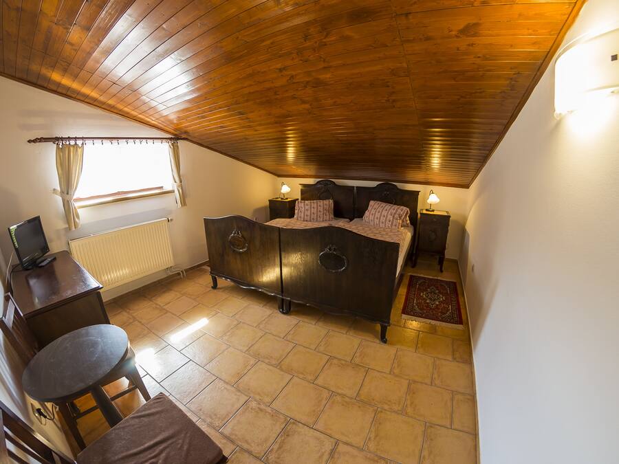 Quarto com teto de madeira, cama com duas almofadas, mesa de cabeceira com abajur, escrivaninha com computador e janela com cortinas.