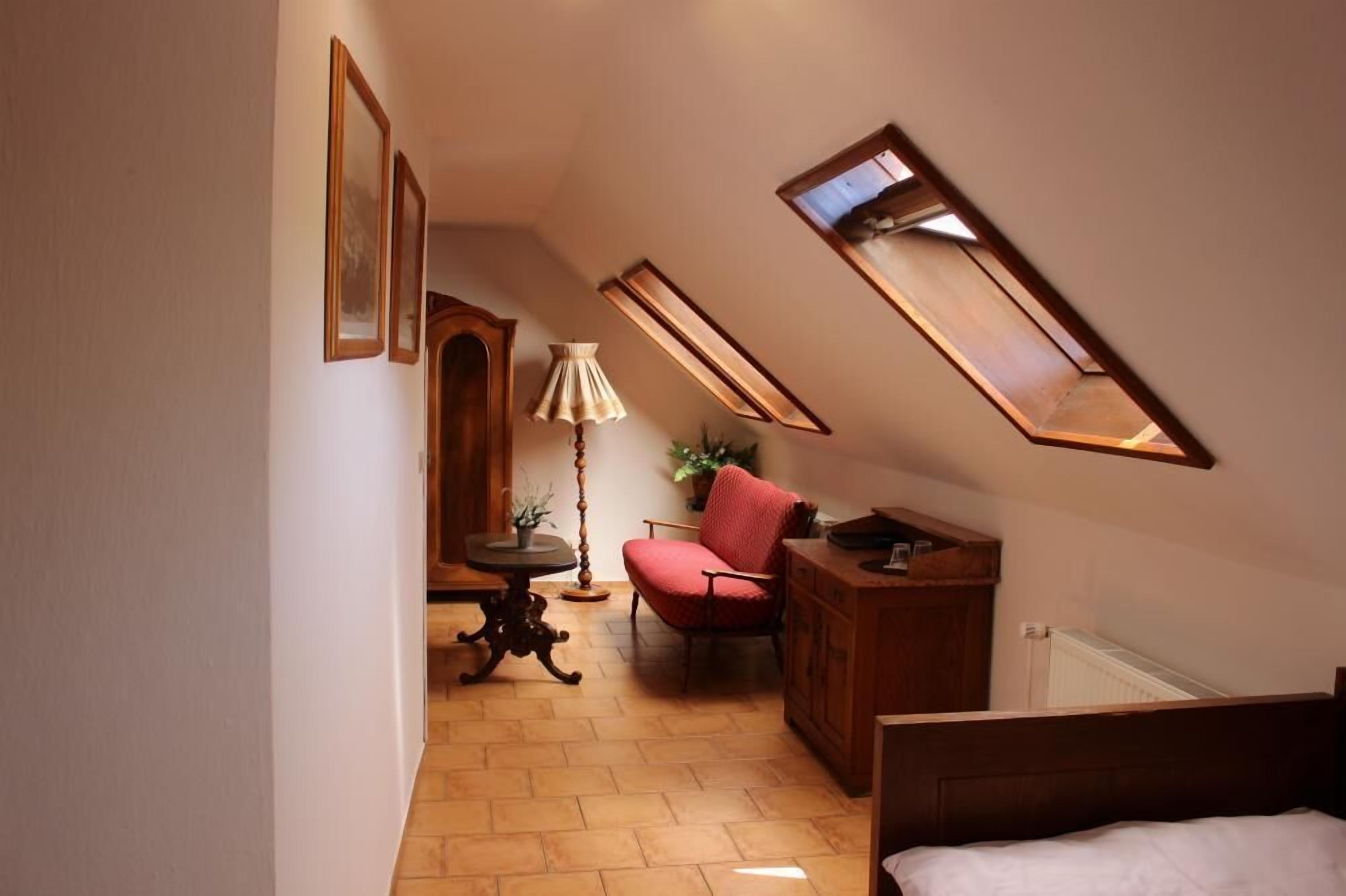Um quarto de sótão acolhedor, com tetto inclinado, uma cama de madeira, uma cadeira vermelha, uma mesinha e um abajur.