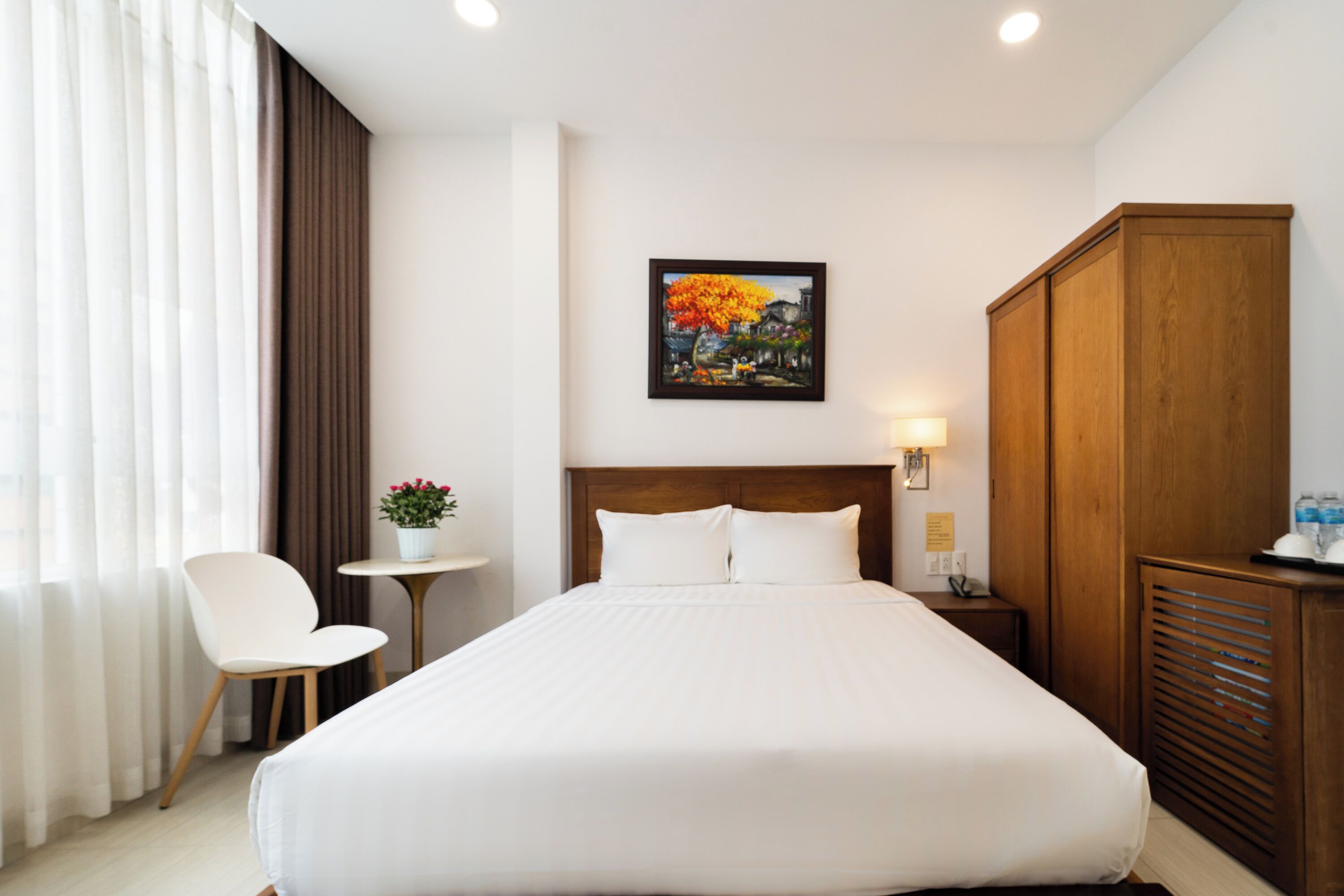Quarto de hotel com uma cama grande, uma mesinha com uma planta em vaso, uma cadeira, um guarda-roupa e um quadro na parede.